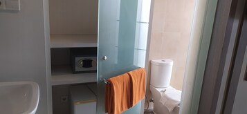 Um banheiro com porta de vidro, vaso sanitário branco e prateleira com rádio.