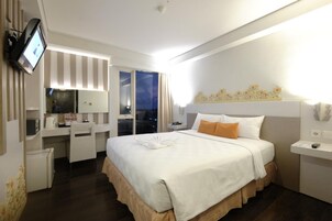 Quarto de hotel com uma cama grande, uma escrivaninha com cadeira, uma televisão e uma janela com cortinas.