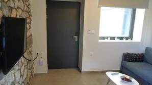 Uma sala de estar moderna com parede de pedra, televisão de tela plana fixada nela, porta escura, janela com persianas, sofá cinza e uma pequena mesa com uma tigela de frutas.