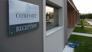 Placa de receção para 'COMFORT SUITES & ROOMS', com fachada de um edifício moderno e uma janela com vista para uma área de estacionamento.