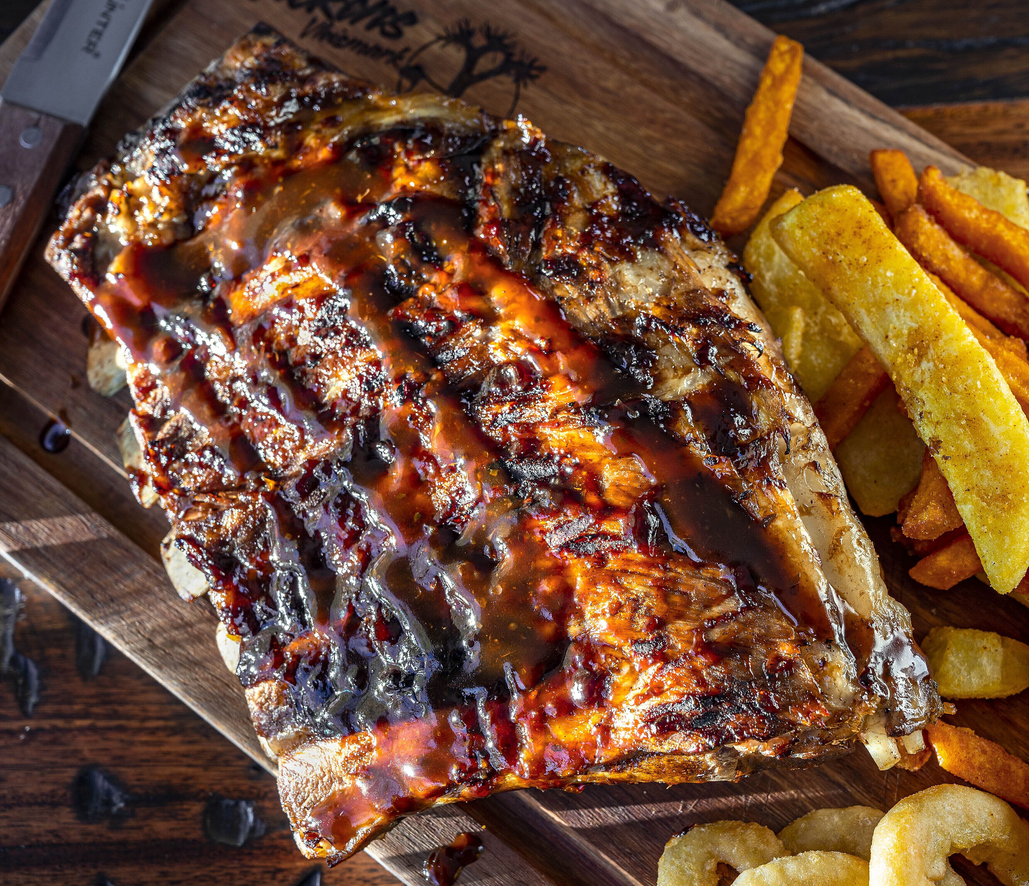 Uma costela de porco grelhada com molho barbecue, acompanhada de batatas fritas e cebolas empanadas em uma tábua de madeira.