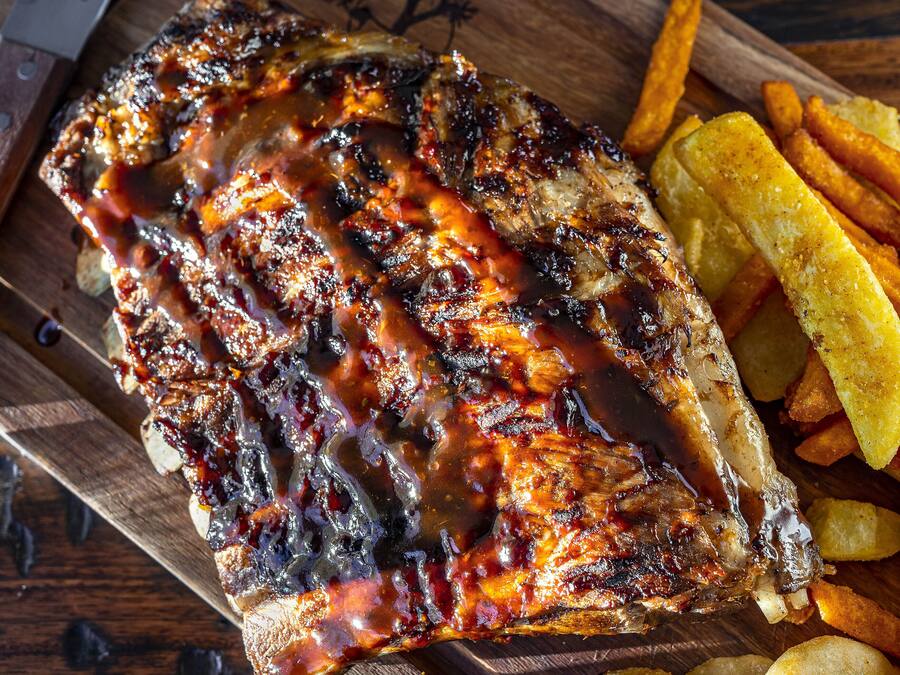 Uma costela de porco grelhada com molho barbecue, acompanhada de batatas fritas e cebolas empanadas em uma tábua de madeira.