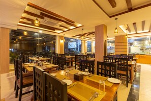Um restaurante com várias mesas e cadeiras, vigas de madeira no teto, luminárias pendentes e janelas amplas.