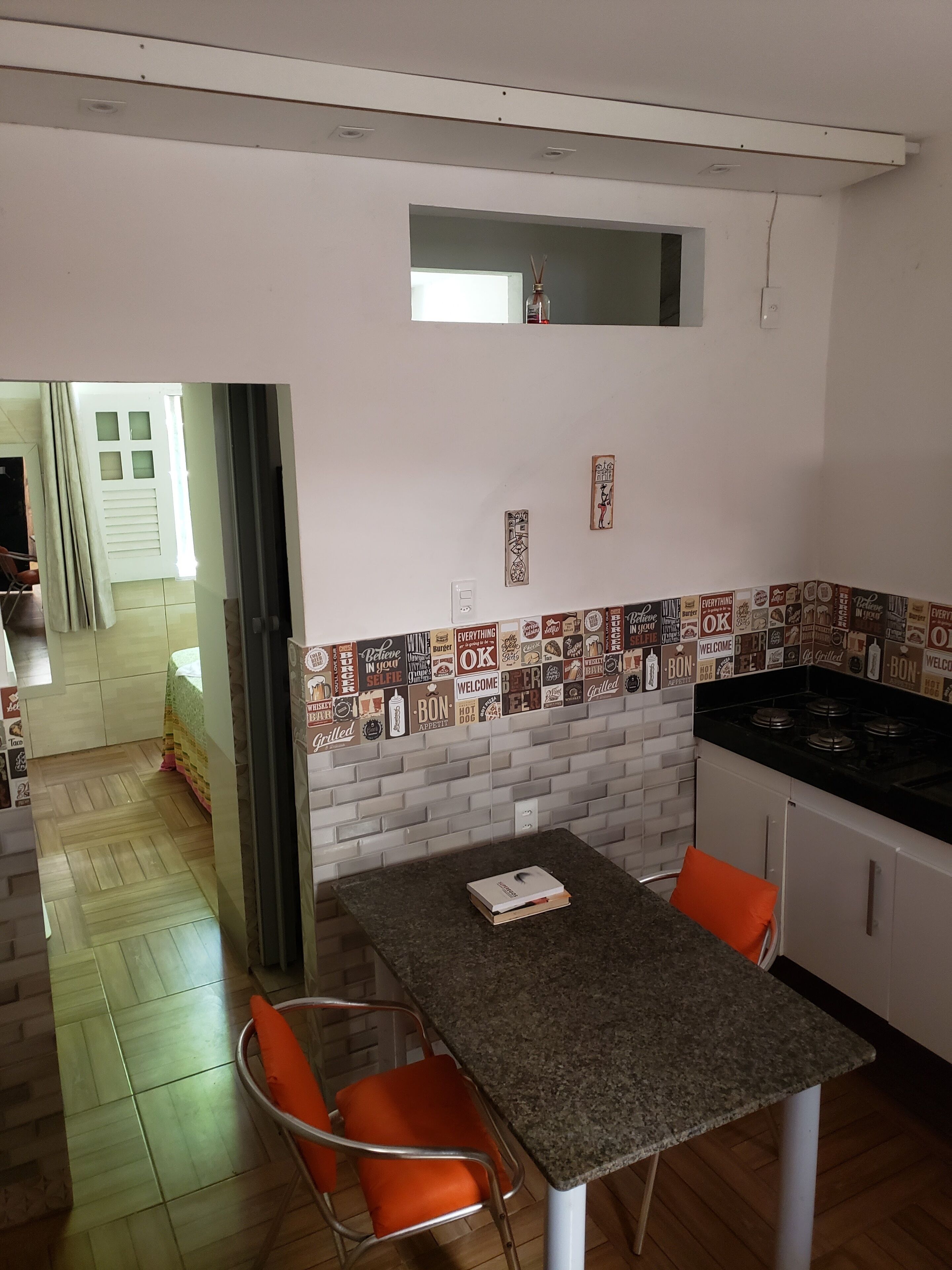 Uma cozinha compacta com área de refeições, com bancada de granito, duas cadeiras laranja, uma pequena janela e painel de parede com padrão de tijolos.