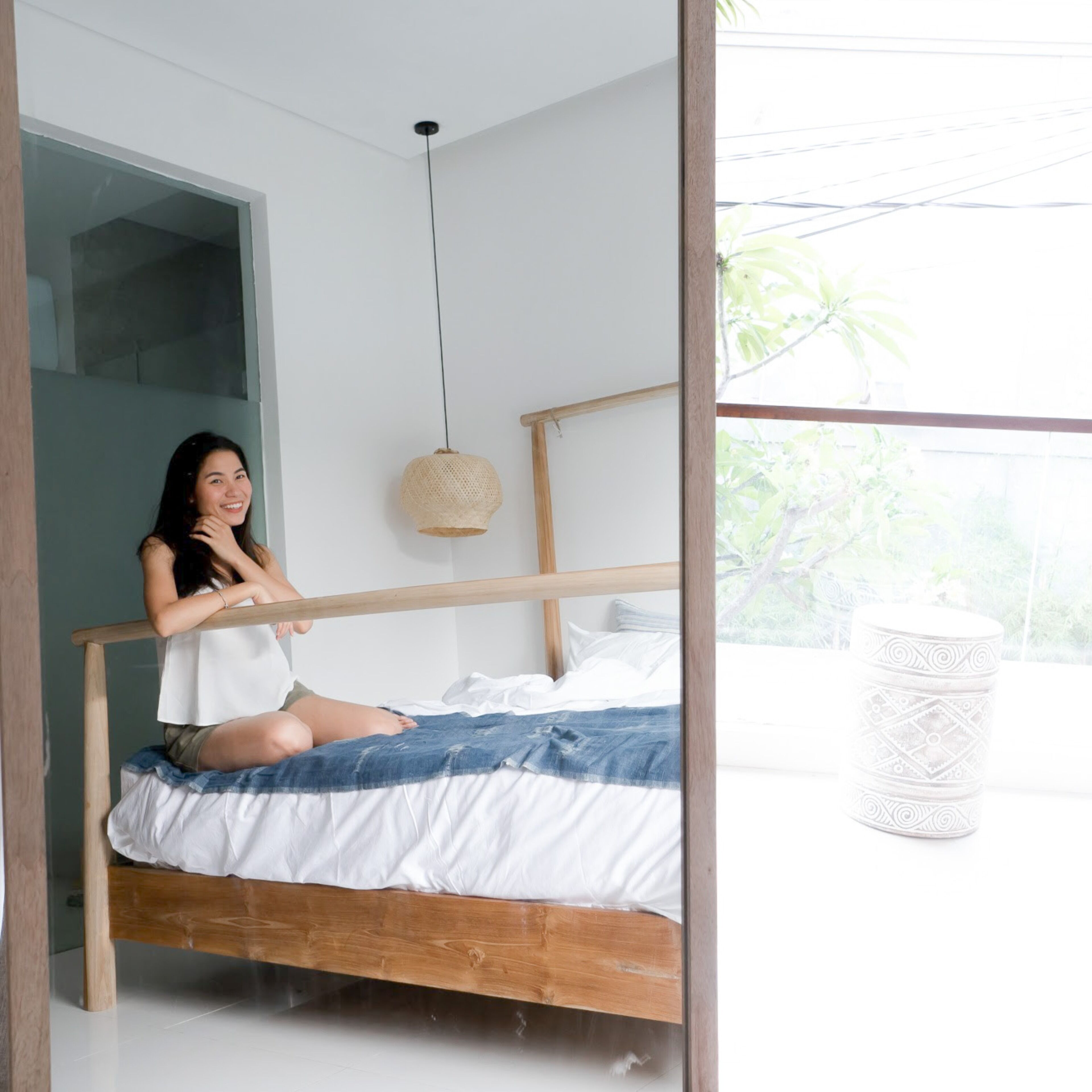 Quarto com cama de madeira, roupa de cama branca, luminária pendente de palha e vista para o verde lá fora.
