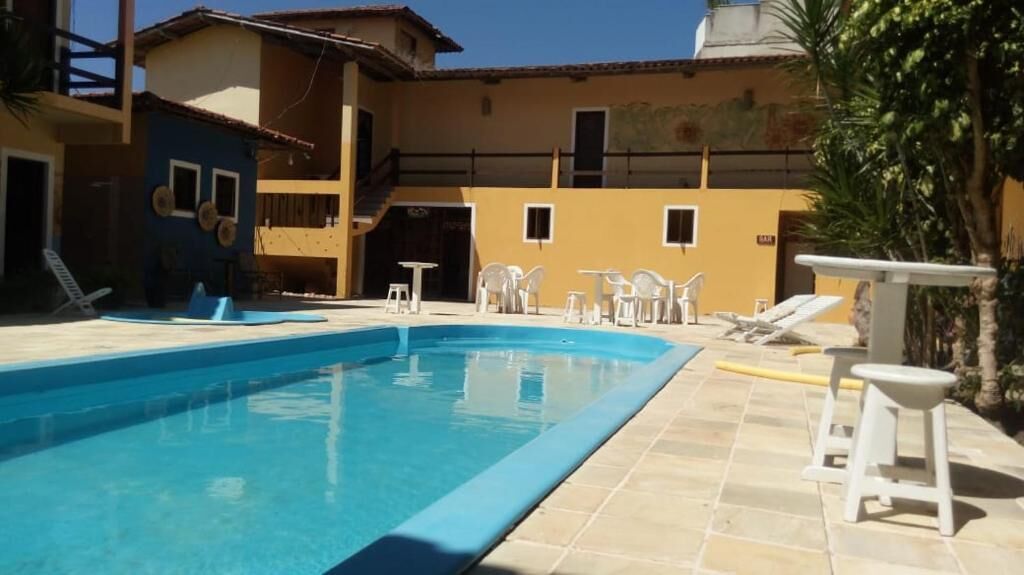 Uma piscina com espreguiçadeiras e mesas, uma escadaria e um edifício com fachada amarela.