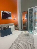 Quarto moderno com cama, almofadas, mesa de cabeceira, espelho e banheiro com vaso sanitário e chuveiro.