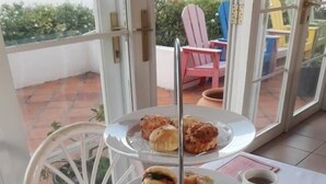 Suporte de sobremesas em três níveis com doces e um bule de chá sobre uma mesa, posicionado numa varanda com cadeiras e vista para vegetação.