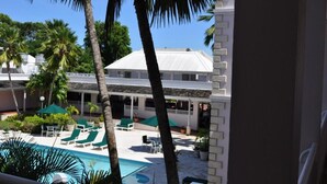 Uma área de piscina com espreguiçadeiras e guarda-sóis, um edifício com fachada branca e telhado vermelho, e palmeiras.