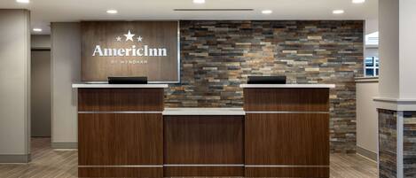 Uma mesa de recepção com acabamento em madeira, uma parede de pedra e uma placa com a inscrição 'American Inn by Wyndham'.
