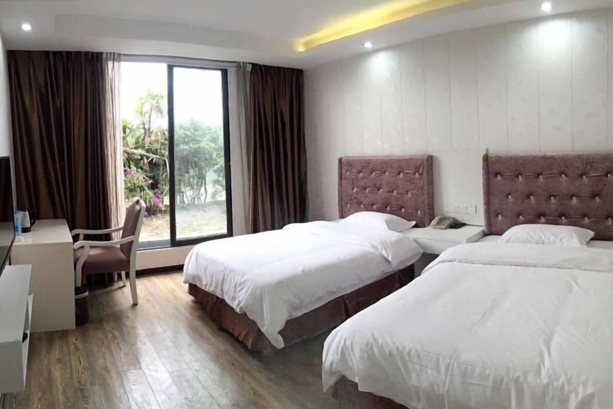 Quarto de hotel com duas camas, uma escrivaninha, uma cadeira e uma janela grande com cortinas.