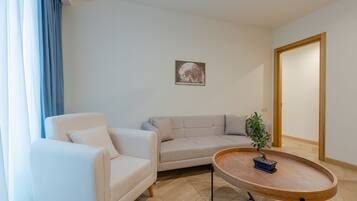 Uma sala de estar moderna com um sofá bege, uma poltrona branca, uma mesa de centro redonda com um bonsai e uma moldura na parede.