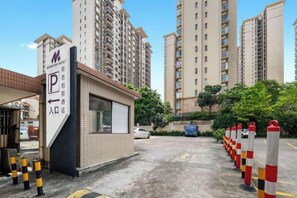 Entrada de hotel com placa de estacionamento, um pequeno posto de recepção e edifícios residenciais altos ao fundo.