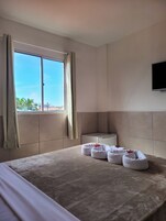 Quarto de hotel com uma cama grande, televisão de tela plana na parede, janela com cortinas e vista para palmeiras e edifícios.