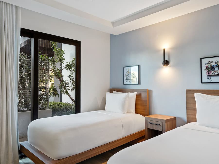Quarto de hotel com duas camas, uma janela com cortinas, uma mesa de cabeceira e obras de arte na parede.