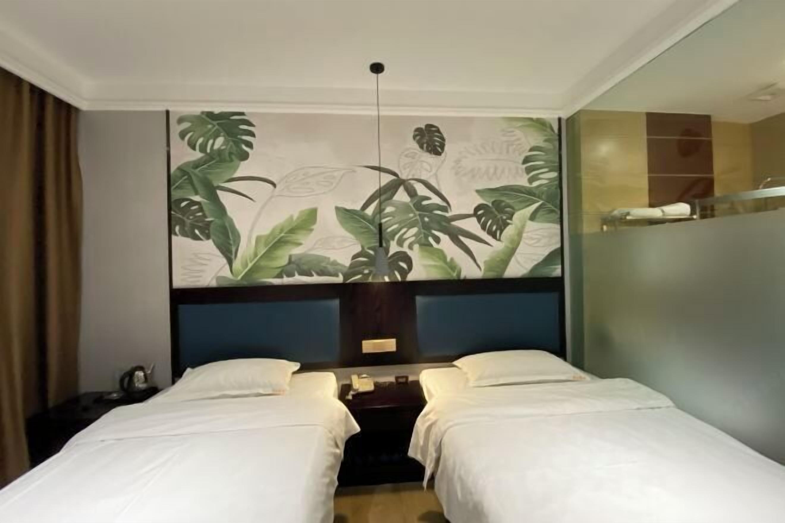Quarto de hotel com duas camas, um grande mural na parede com folhas tropicais, uma cabeceira moderna e uma mesa de cabeceira com abajur.