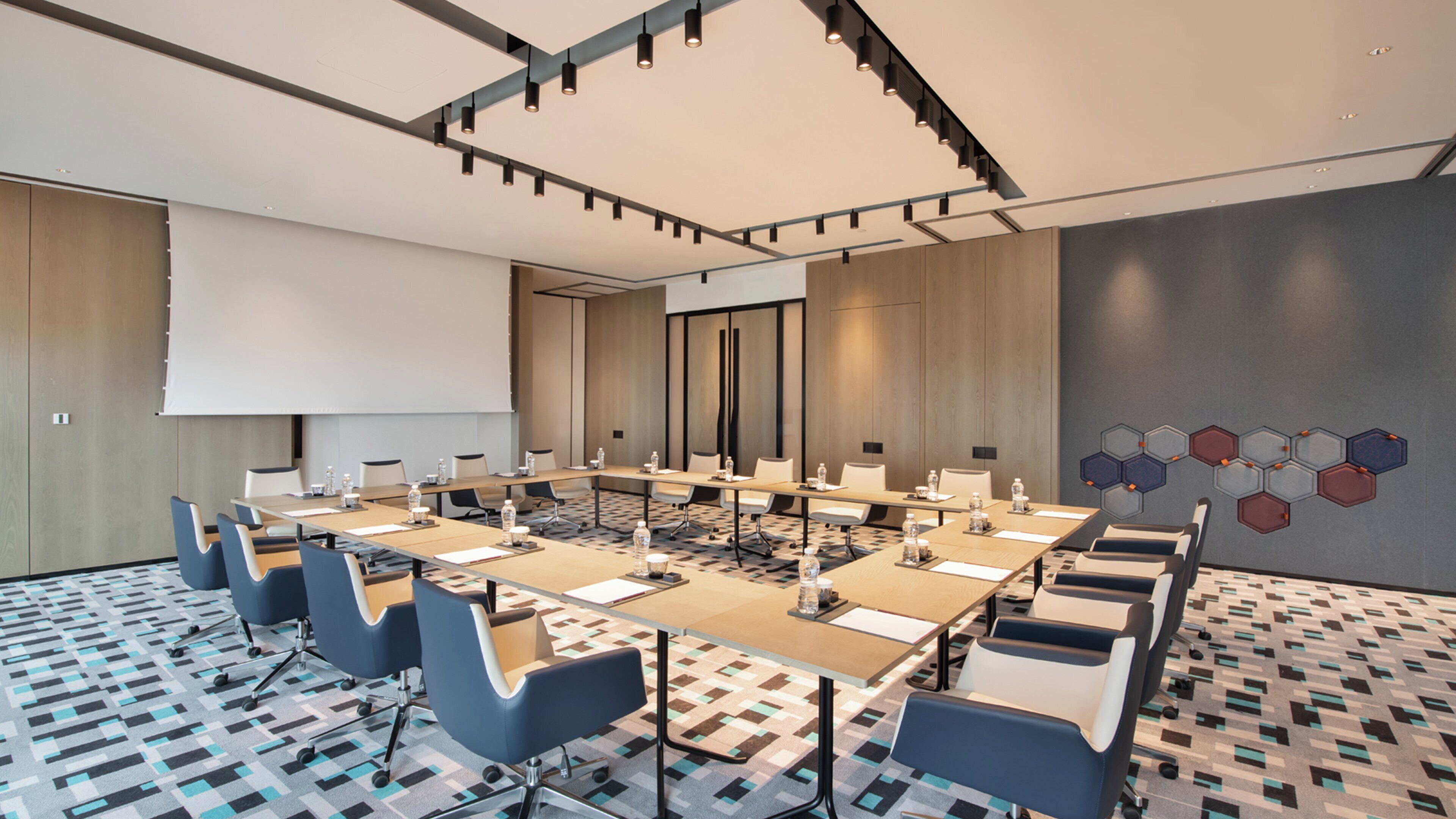 Uma sala de conferências com uma grande mesa, cadeiras, uma tela de projetor e uma decoração de parede hexagonal.