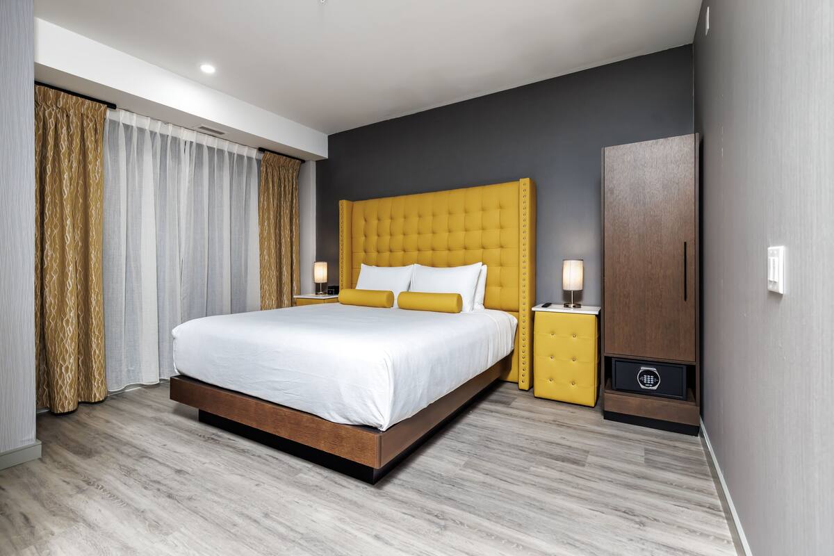 Quarto de hotel moderno com uma cama grande, cabeceira de madeira, mesa de cabeceira com luminária e guarda-roupa.