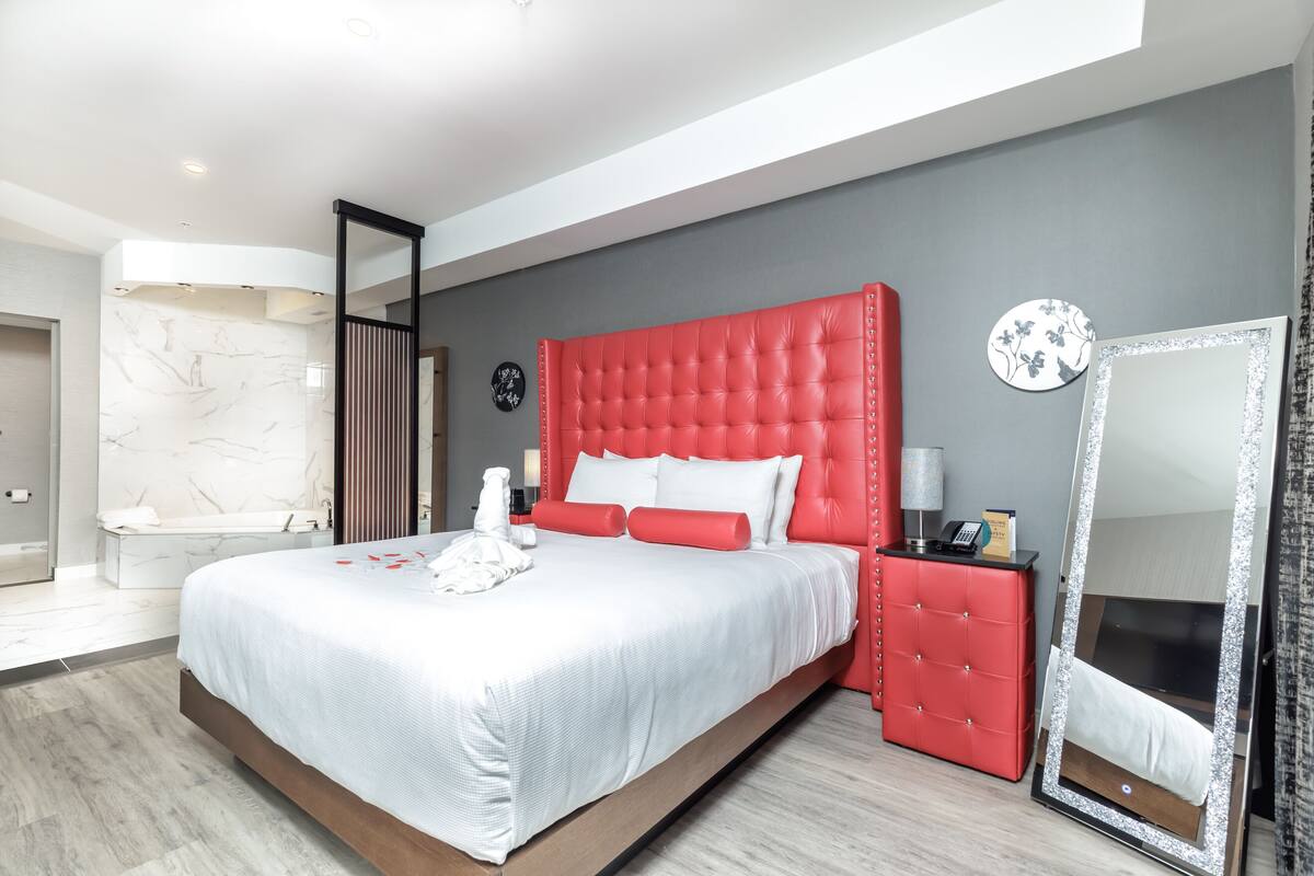 Quarto de hotel moderno com uma cama grande, cabeceira vermelha, mesa de cabeceira com luminária e um espelho amplo.