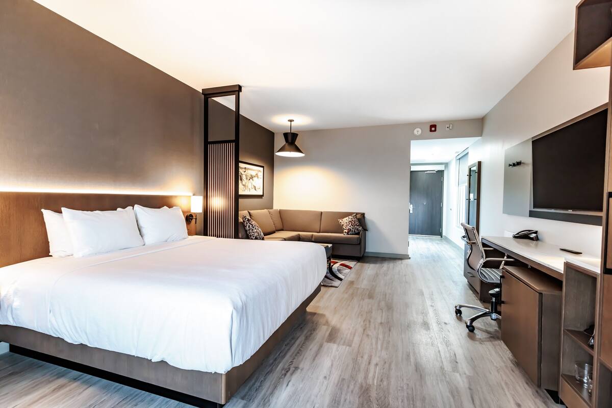 Quarto de hotel moderno com uma cama grande, um sofá, uma escrivaninha com cadeira, televisão e piso de madeira.