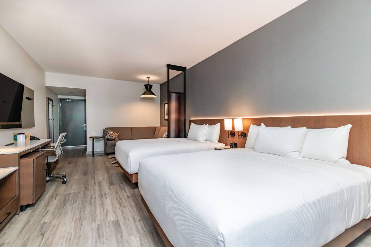 Quarto de hotel com duas camas, uma escrivaninha, uma cadeira, uma TV e uma mesinha de cabeceira com abajur.