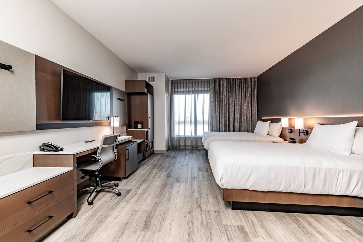 Quarto de hotel moderno com uma cama grande, uma mesa com cadeira, televisão de tela plana e uma janela ampla com cortinas.