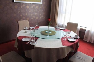 Uma mesa de jantar com tampo de vidro, rodeada por cadeiras, posta com pratos, copos e um vaso com uma rosa.