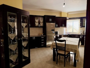 Uma cozinha com armários de madeira escura, geladeira de aço inoxidável, mesa de jantar com cadeiras e uma janela com cortinas.