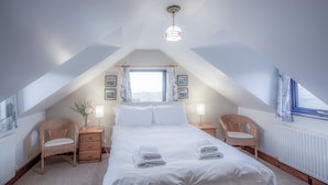 Um quarto com uma cama grande, duas mesas de cabeceira, duas cadeiras de vime, uma janela com cortinas e quadros nas paredes.