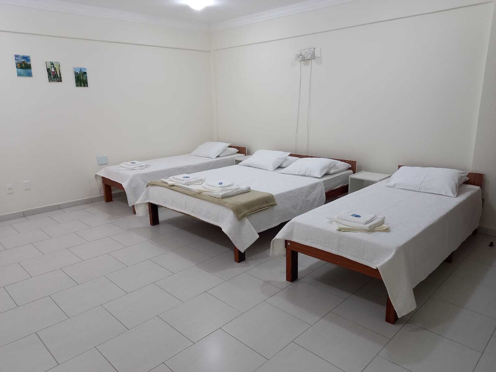 Quarto com três camas de solteiro, roupa de cama branca e chão em azulejo.