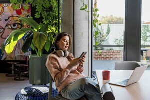 Uma pessoa sentada a uma mesa com um laptop, fones de ouvido e um smartphone.