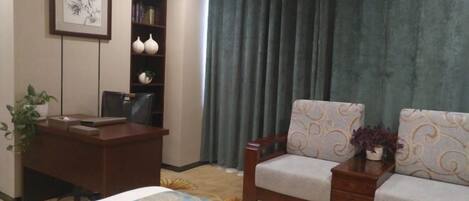 Quarto de hotel com escrivaninha, duas cadeiras, estante de livros, janela ampla com cortinas e cama com colcha estampada.