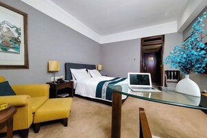 Quarto de hotel com cama, escrivaninha com laptop, poltrona amarela, mesa de vidro e planta decorativa.
