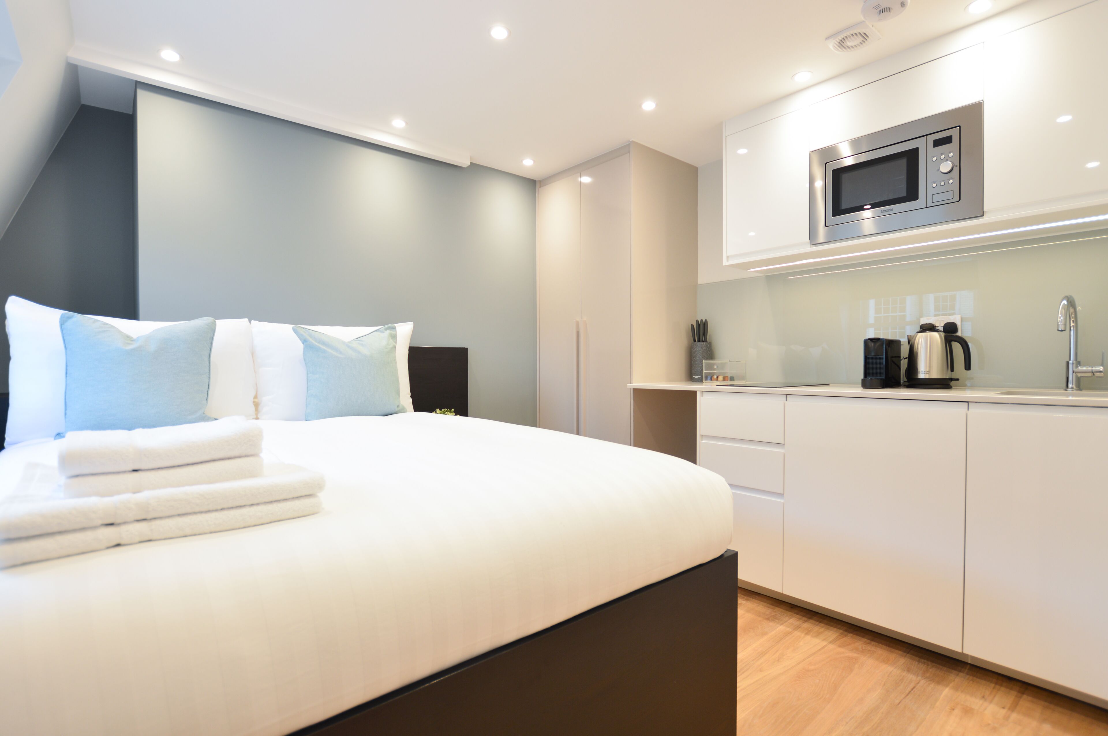Quarto de hotel moderno com cama bem arrumada, cozinha compacta com micro-ondas e bancada pequena.