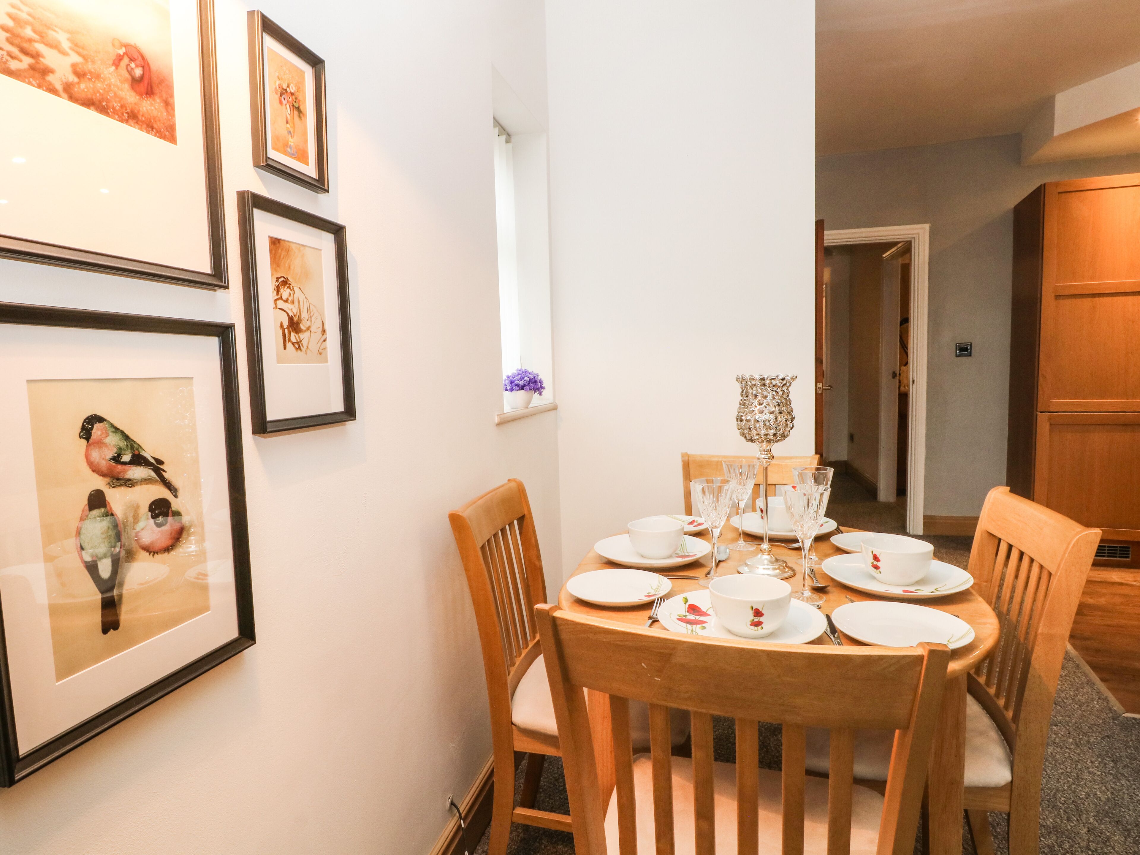 Uma área de refeições com mesa de madeira posta para quatro pessoas, arte decorativa na parede e uma janela com um vaso de flores.