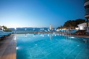 Uma piscina com espreguiçadeiras e guarda-sóis, vista para o mar e um prédio com varanda.