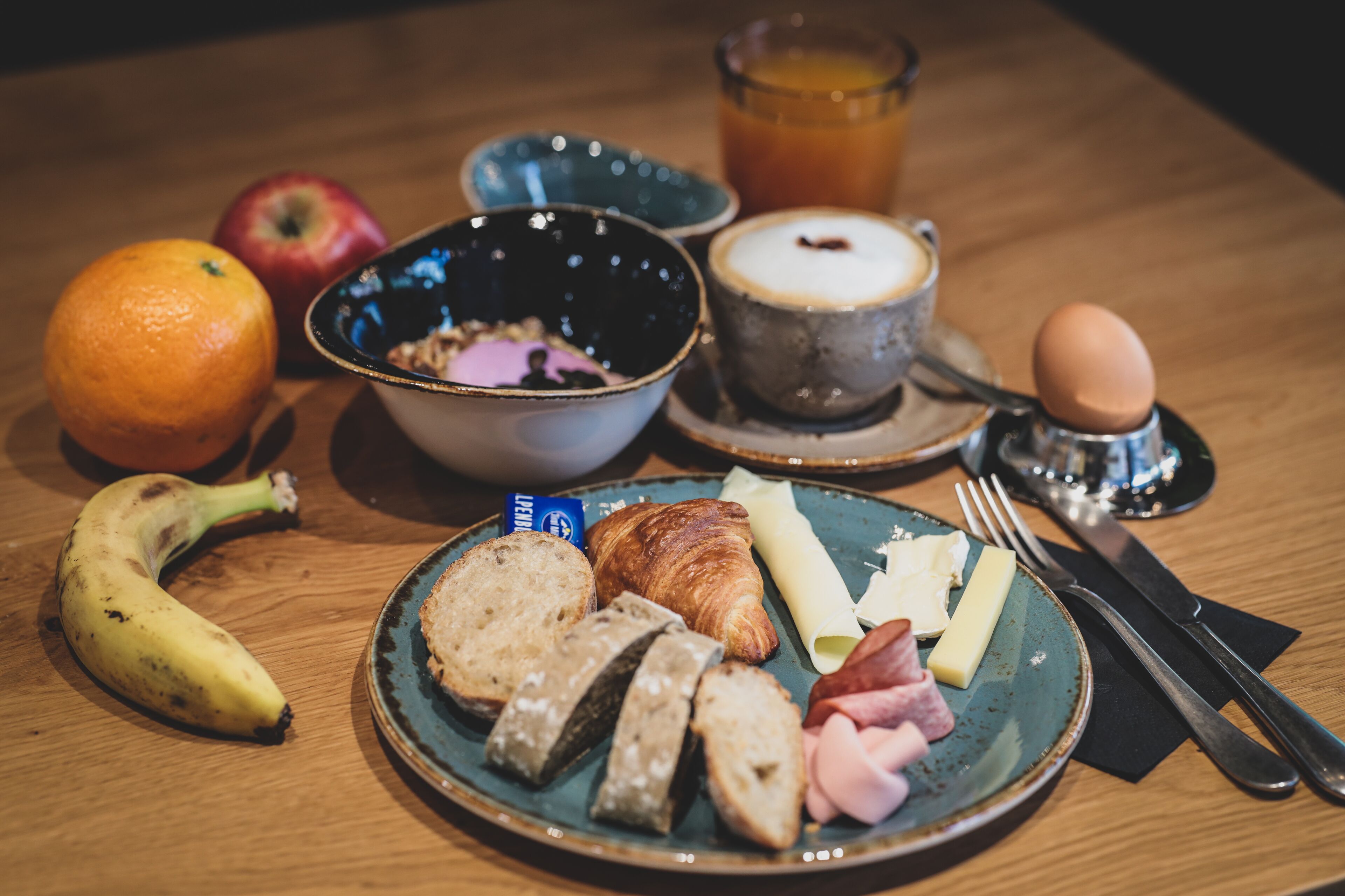 Uma mesa de café da manhã com uma travessa de croissants, queijo, presunto e pão, uma tigela de cereal, uma laranja, uma maçã, um copo de suco de laranja e uma xícara de café.