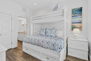 Uma cama beliche com um cobreleito estampado, uma mesinha de cabeceira com abajur e uma parede com duas pinturas emolduradas.