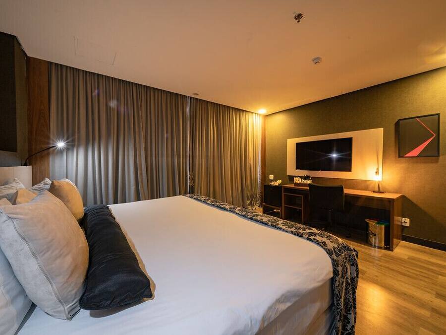 Quarto de hotel com uma cama grande, uma escrivaninha, uma televisão e uma cortina.