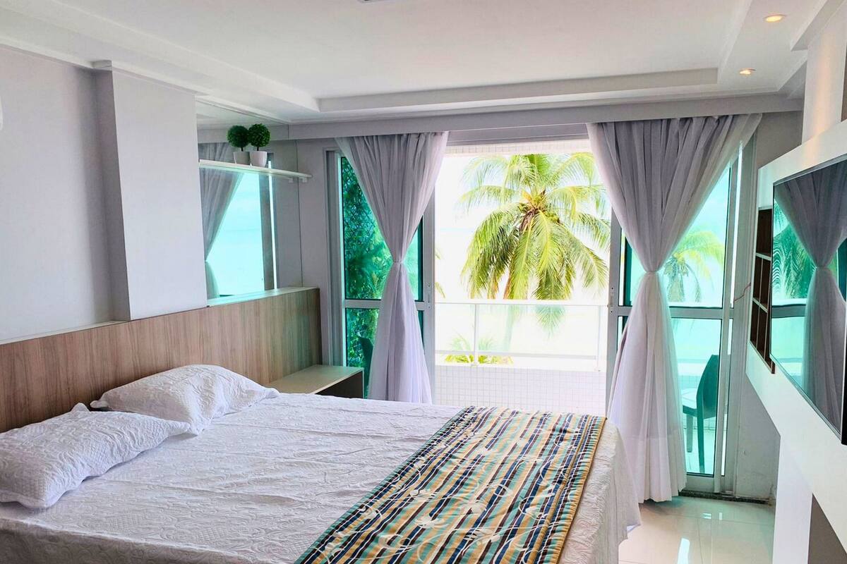 Quarto de hotel moderno com uma cama grande, uma televisão e varanda com vista para palmeiras.