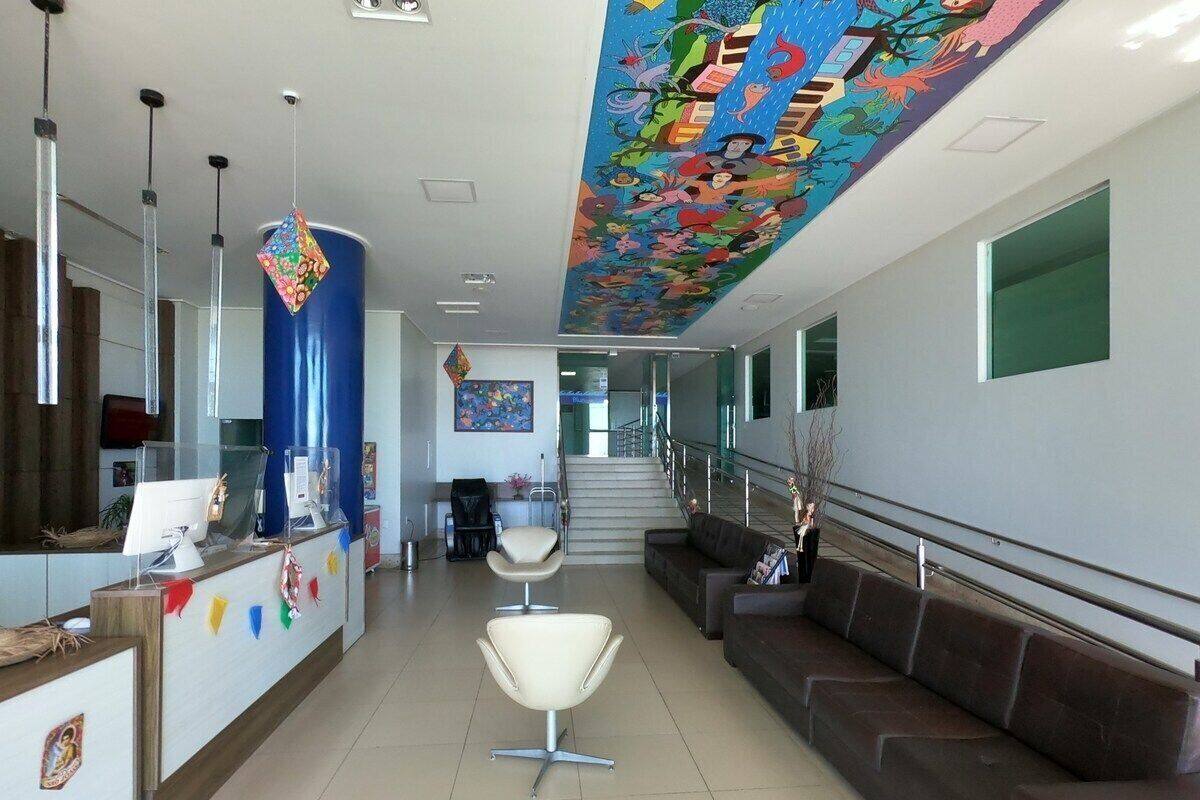 Uma área de recepção com balcão, duas cadeiras brancas, um sofá castanho e um mural colorido no teto.