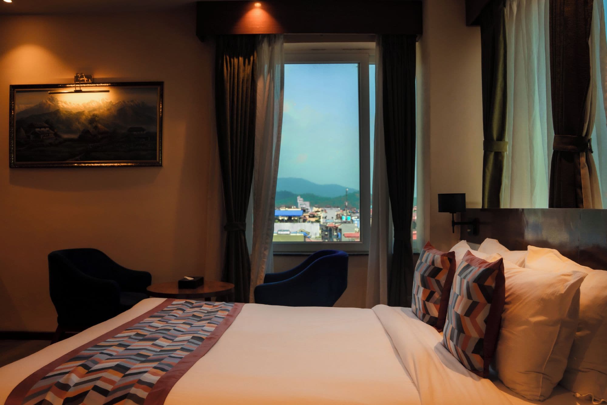 Quarto de hotel com cama, escrivaninha, cadeira, janela com vista para a cidade e um quadro na parede.