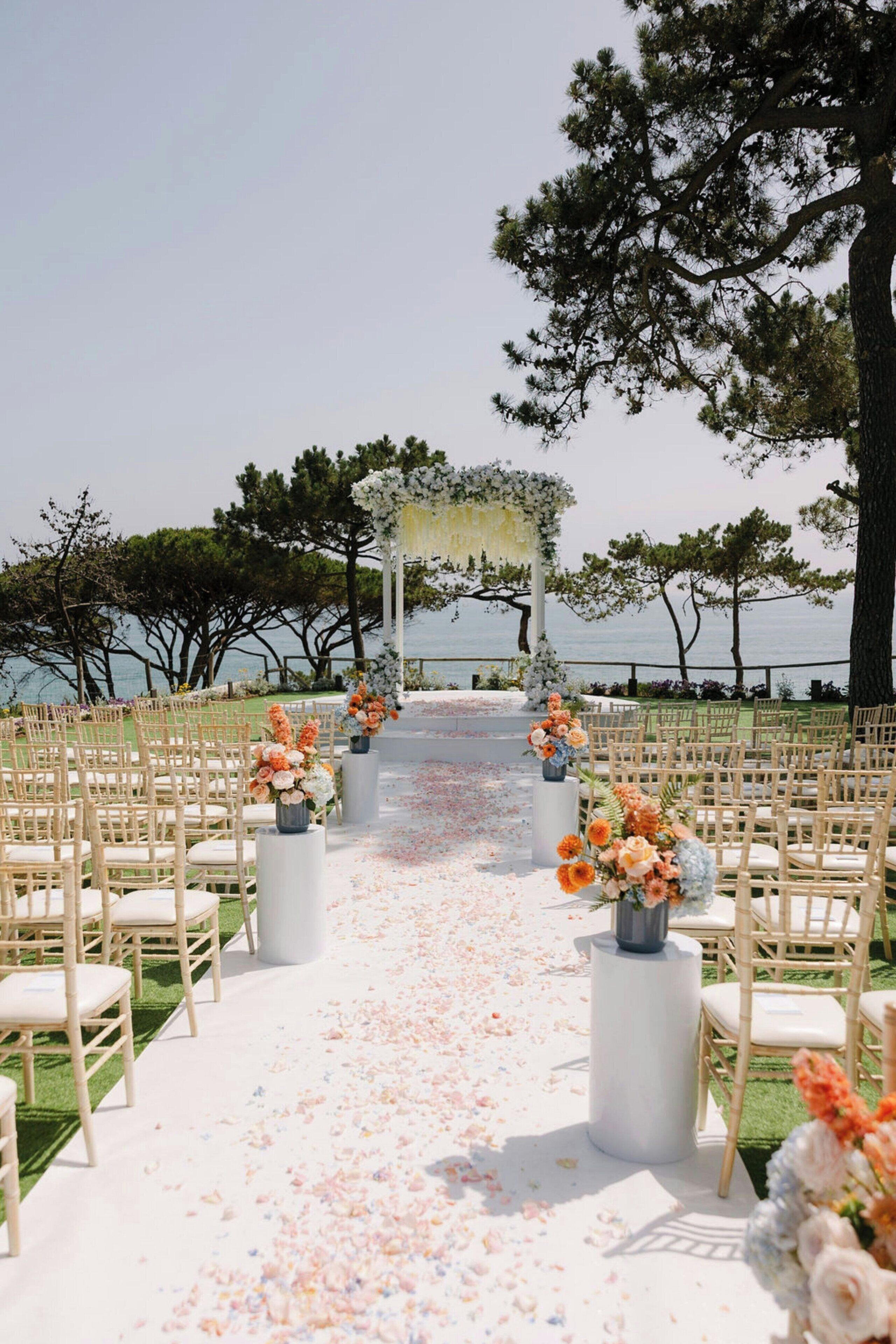 Um corredor de casamento com cadeiras brancas e arranjos florais, levando a um arco branco.