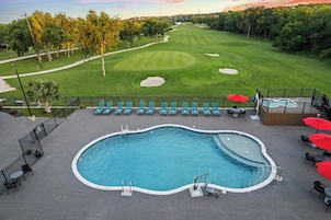 Uma piscina com espreguiçadeiras e guarda-sóis, cercada por uma área ajardinada com árvores e um campo de golfe ao fundo.