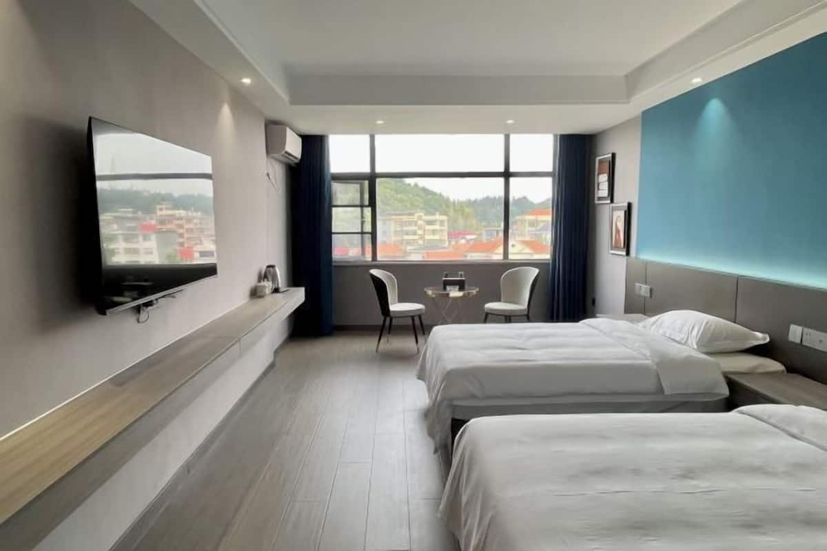 Quarto de hotel com duas camas, televisão, uma pequena mesa com cadeiras e vista para prédios através de amplas janelas.