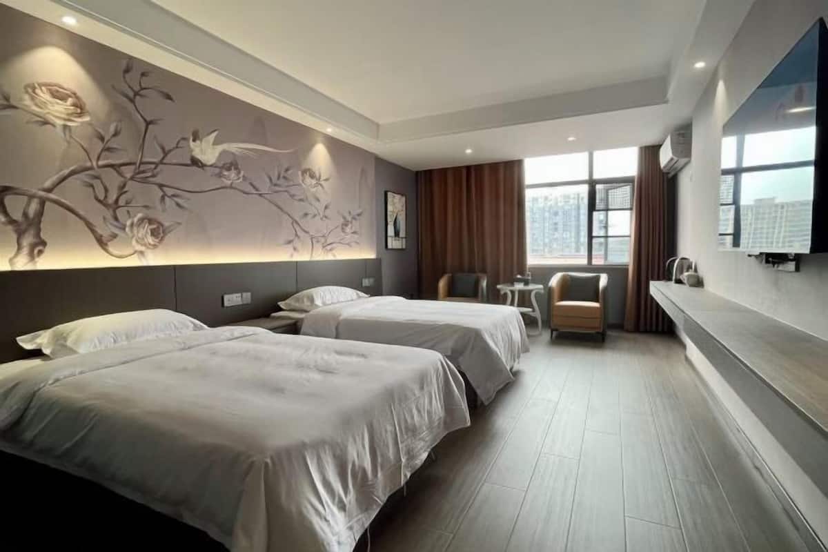 Quarto de hotel com duas camas, uma mesinha, uma cadeira, uma janela grande e uma televisão presa na parede.