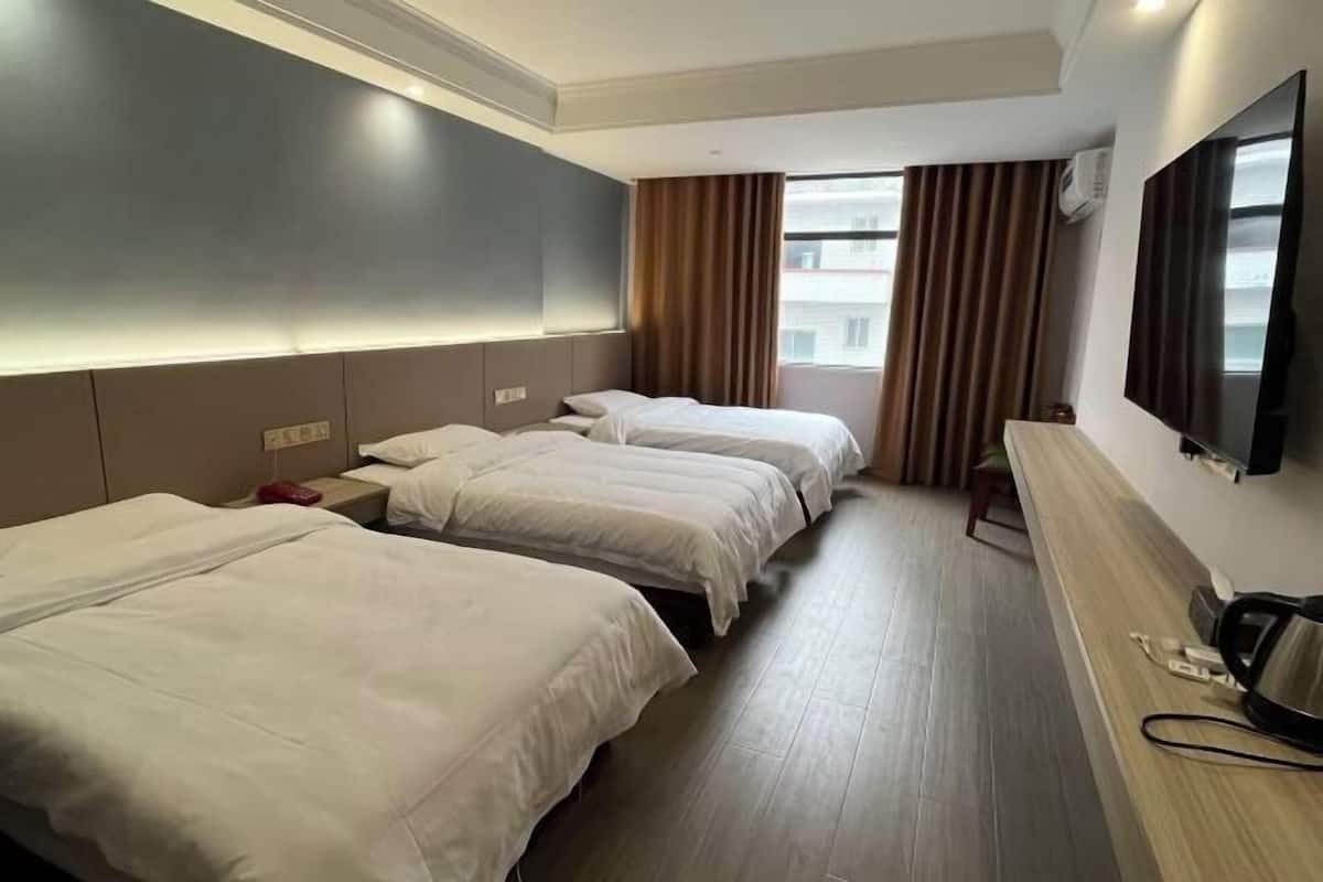 Quarto de hotel com três camas, uma escrivaninha de madeira, televisão de tela plana e uma janela com cortinas.