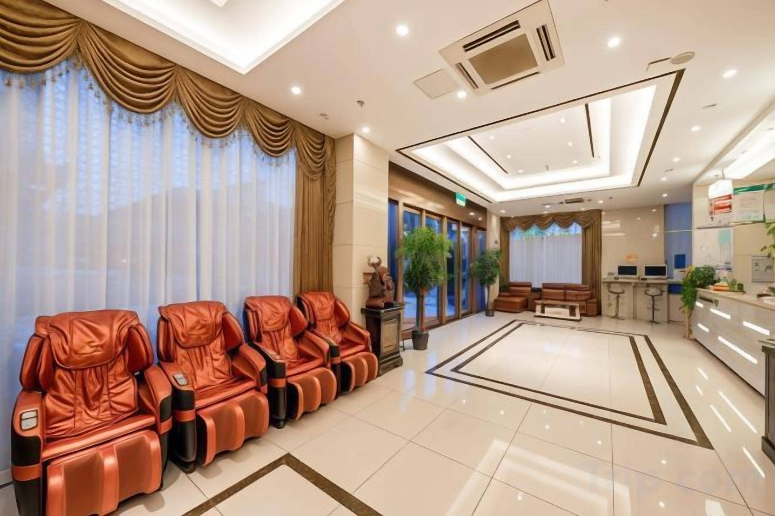 Um saguão de hotel com cadeiras de massagem, balcão de recepção e uma grande janela com cortinas.