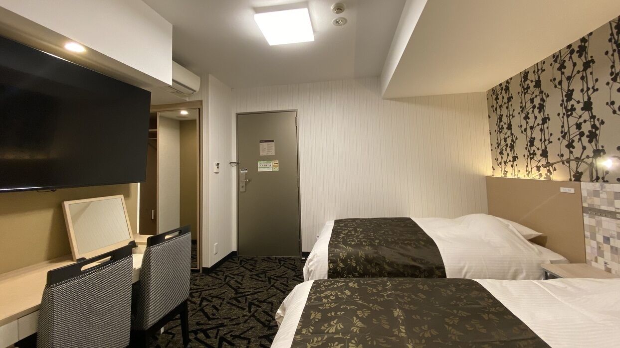 Quarto de hotel com duas camas, uma escrivaninha com cadeira, TV de tela plana e porta para um corredor.