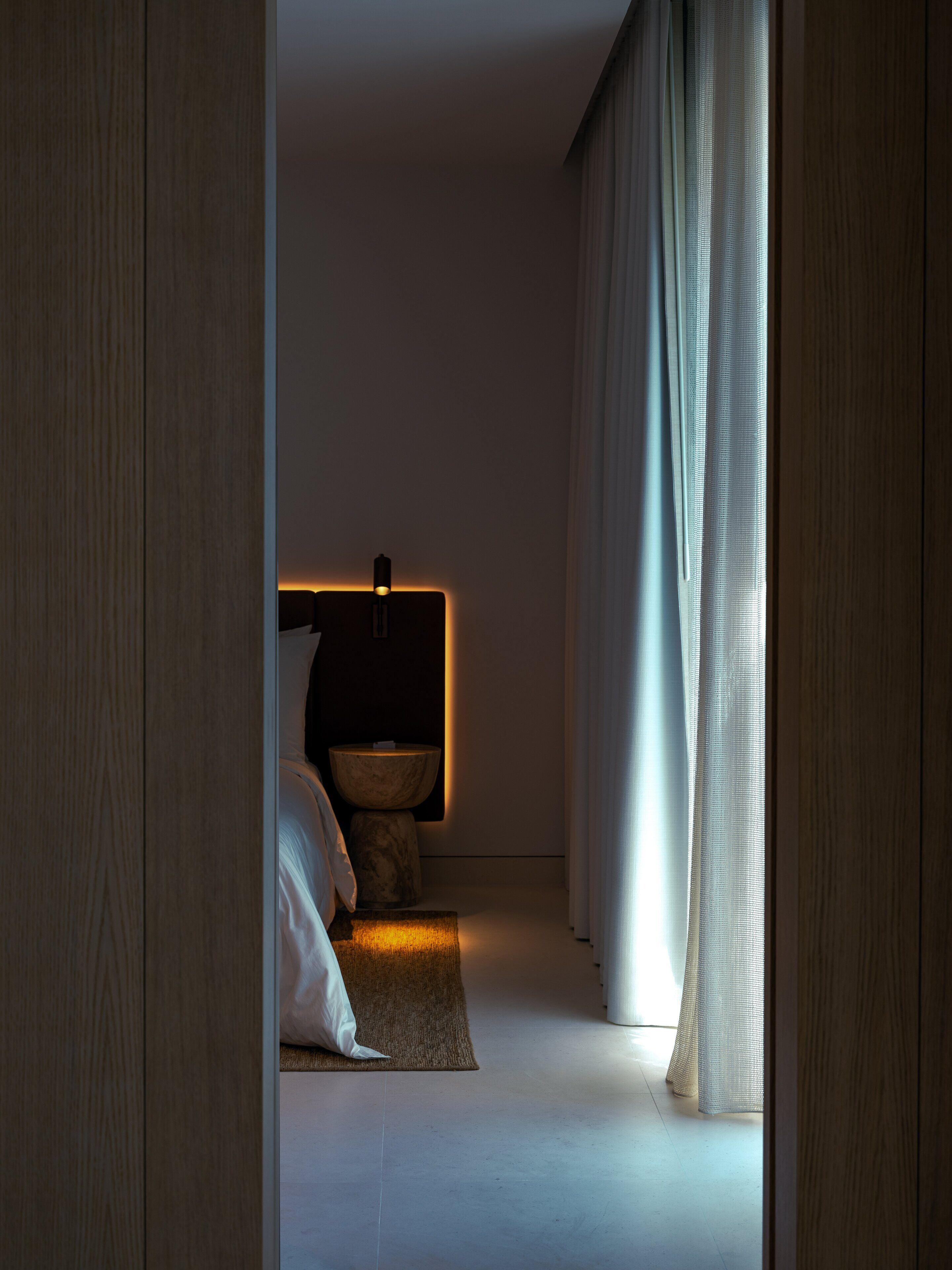 Quarto moderno com design minimalista, apresentando cama com roupa de cama branca, mesa de cabeceira com abajur e uma janela grande com cortinas finas.